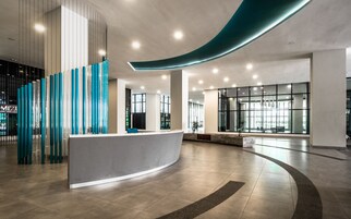 Uma área de recepção moderna com um balcão curvo, um pilar decorativo azul-turquesa e grandes janelas que permitem a entrada de luz natural.