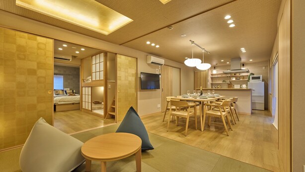 Quarto de hotel moderno com piso de madeira, área de refeições com mesa e cadeiras, cozinha equipada com micro-ondas e geladeira, e TV fixada na parede.
