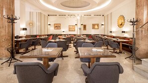 Ampla sala de jantar de hotel, com várias mesas e cadeiras, um candelabro central e obras de arte emolduradas nas paredes.