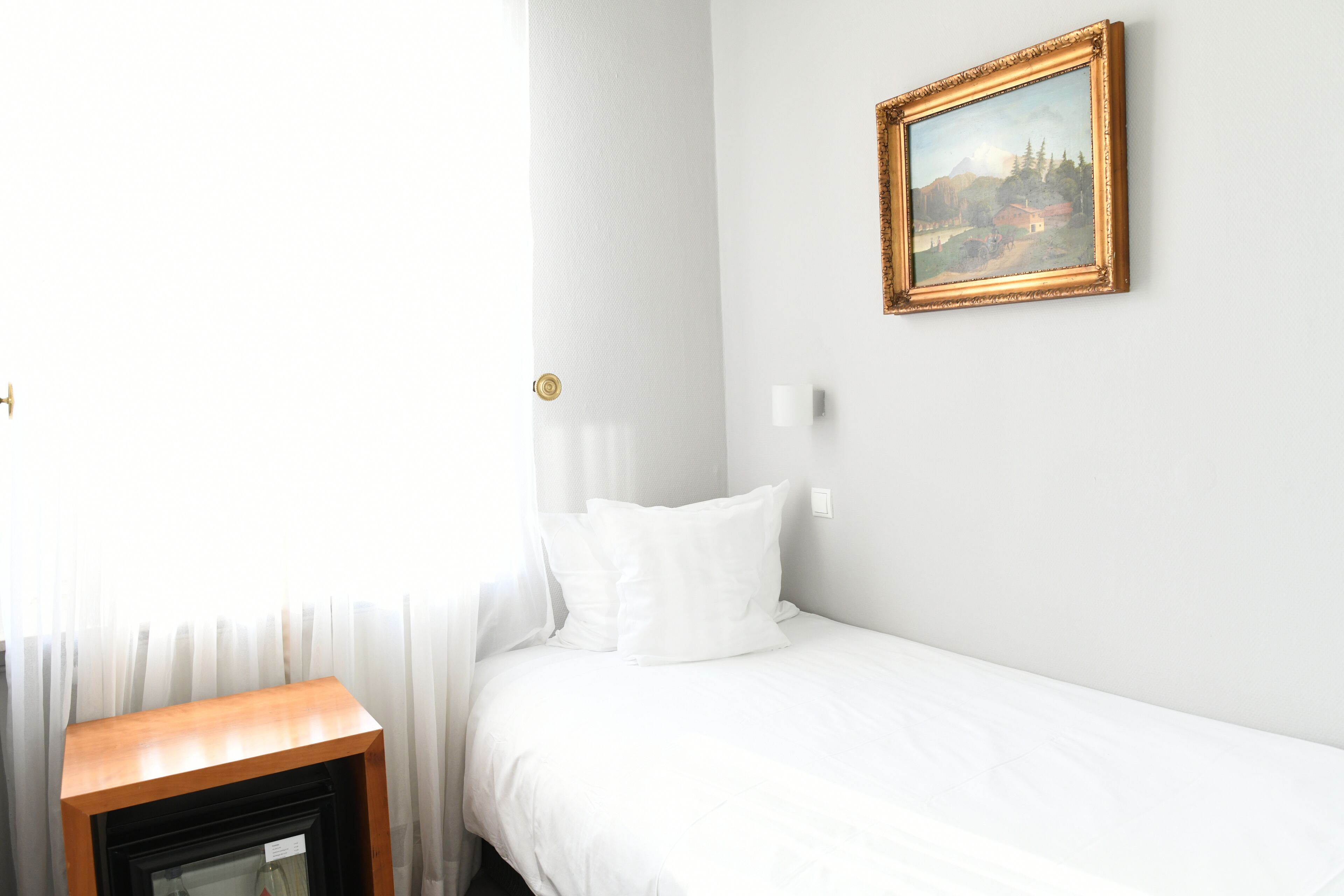 Quarto de hotel bem arrumado, com uma cama, uma pequena mesa de madeira, um quadro na parede e uma janela com cortinas finas.