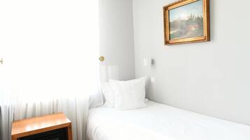 Quarto de hotel bem arrumado, com uma cama, uma pequena mesa de madeira, um quadro na parede e uma janela com cortinas finas.