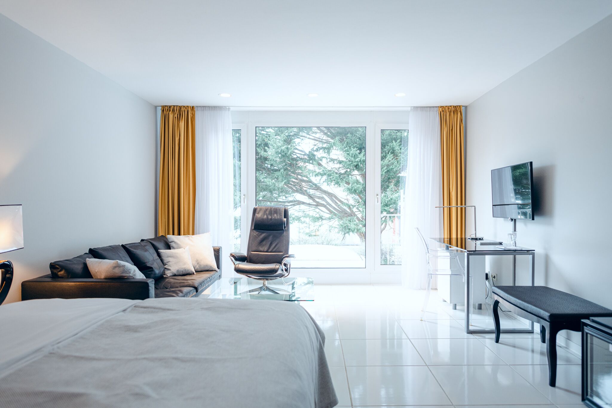 Quarto de hotel moderno com cama, sofá, poltrona, escrivaninha e televisão.