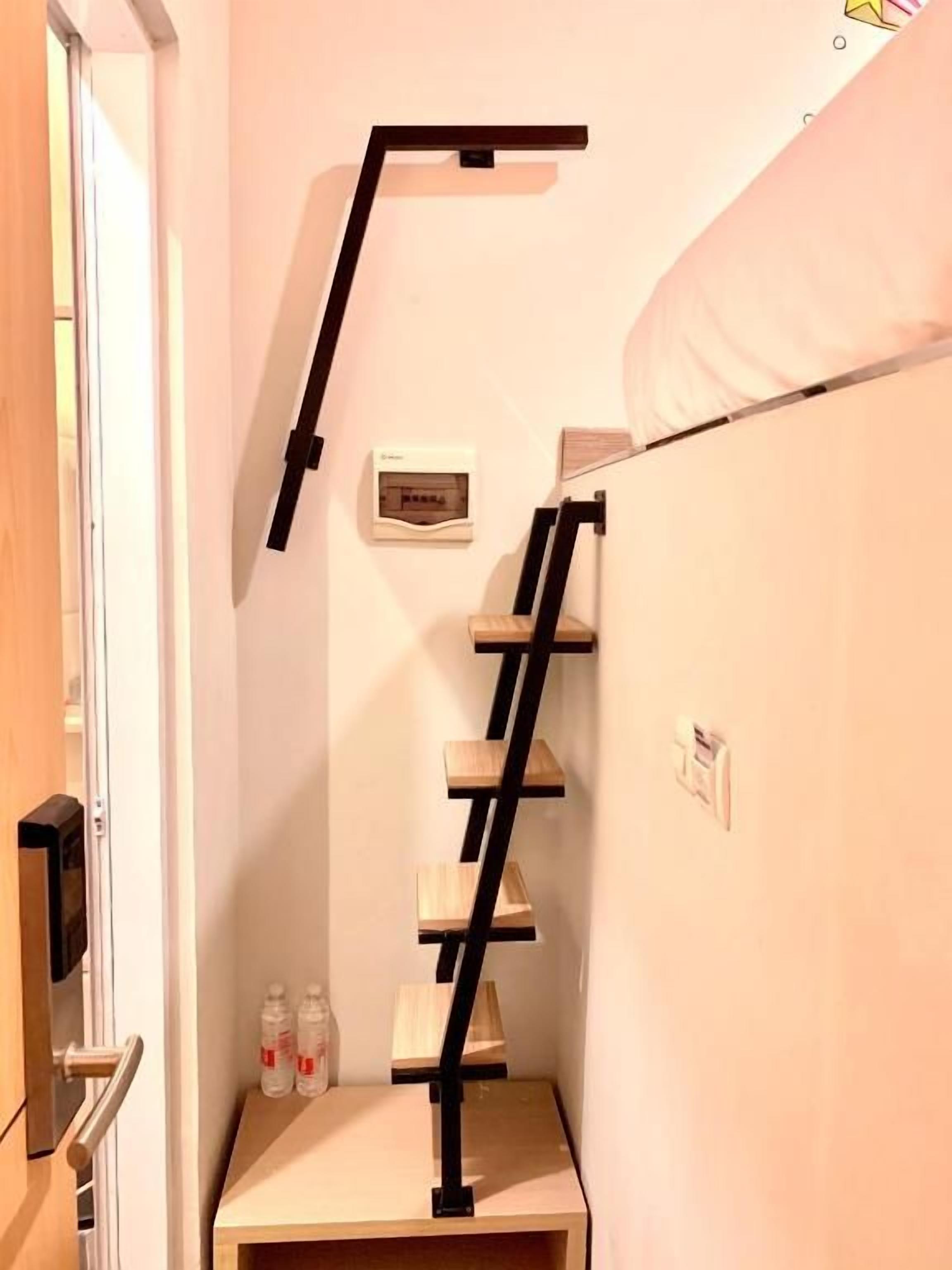 Quarto estreito com estante de madeira e degraus, uma porta e uma pequena mesa com garrafas.