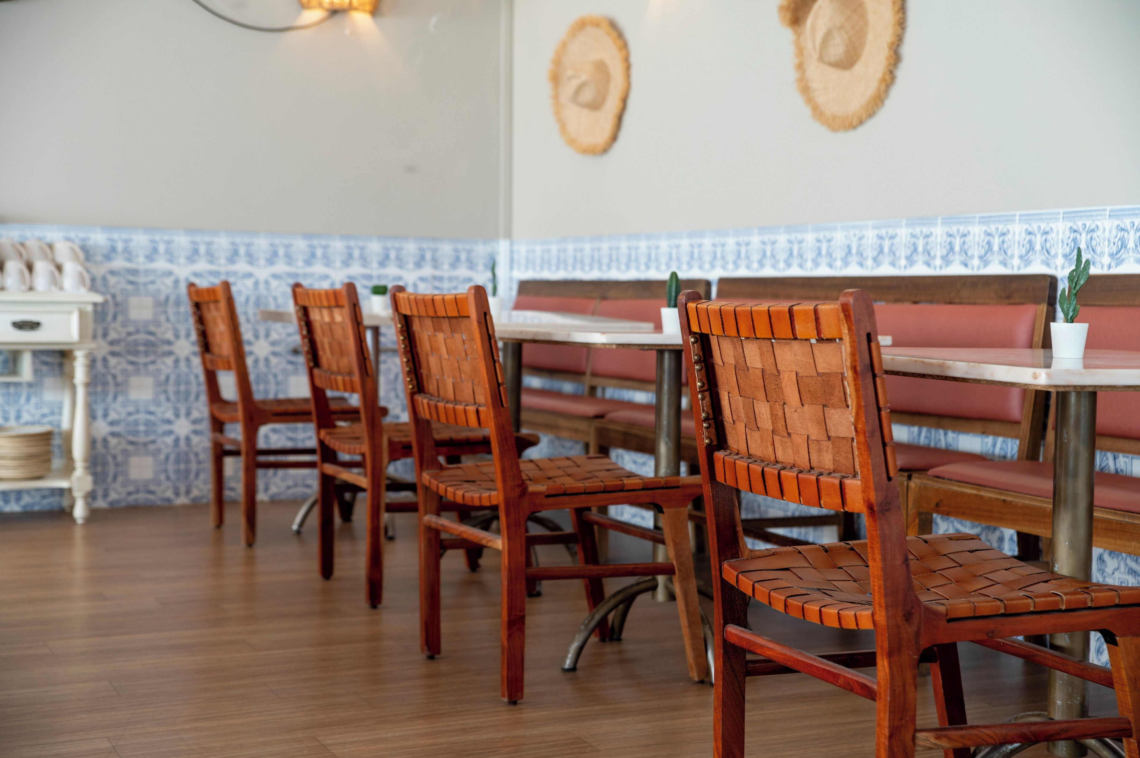 Interior de restaurante com cadeiras e mesas de madeira, azulejos azuis e brancos nas paredes e uma estante com objetos à esquerda.