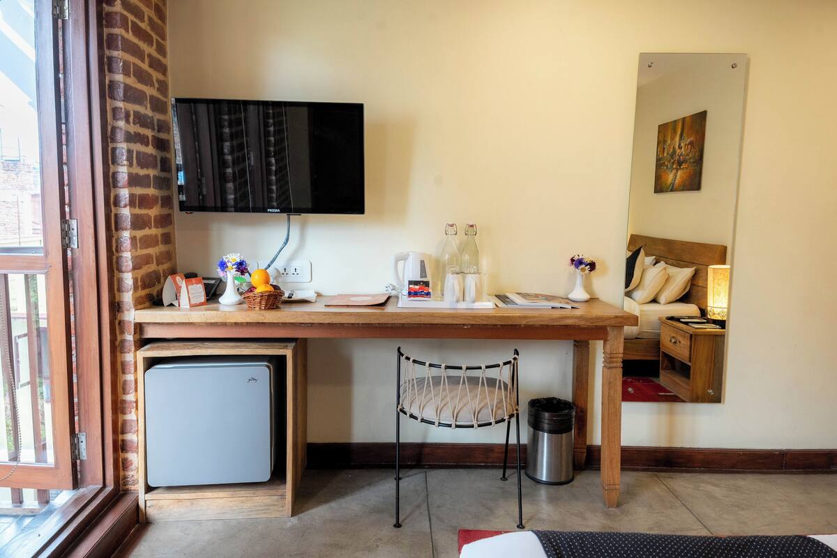 Quarto compacto com escrivaninha, minibar, cadeira, cama com almofadas, mesa de cabeceira com abajur e TV na parede.