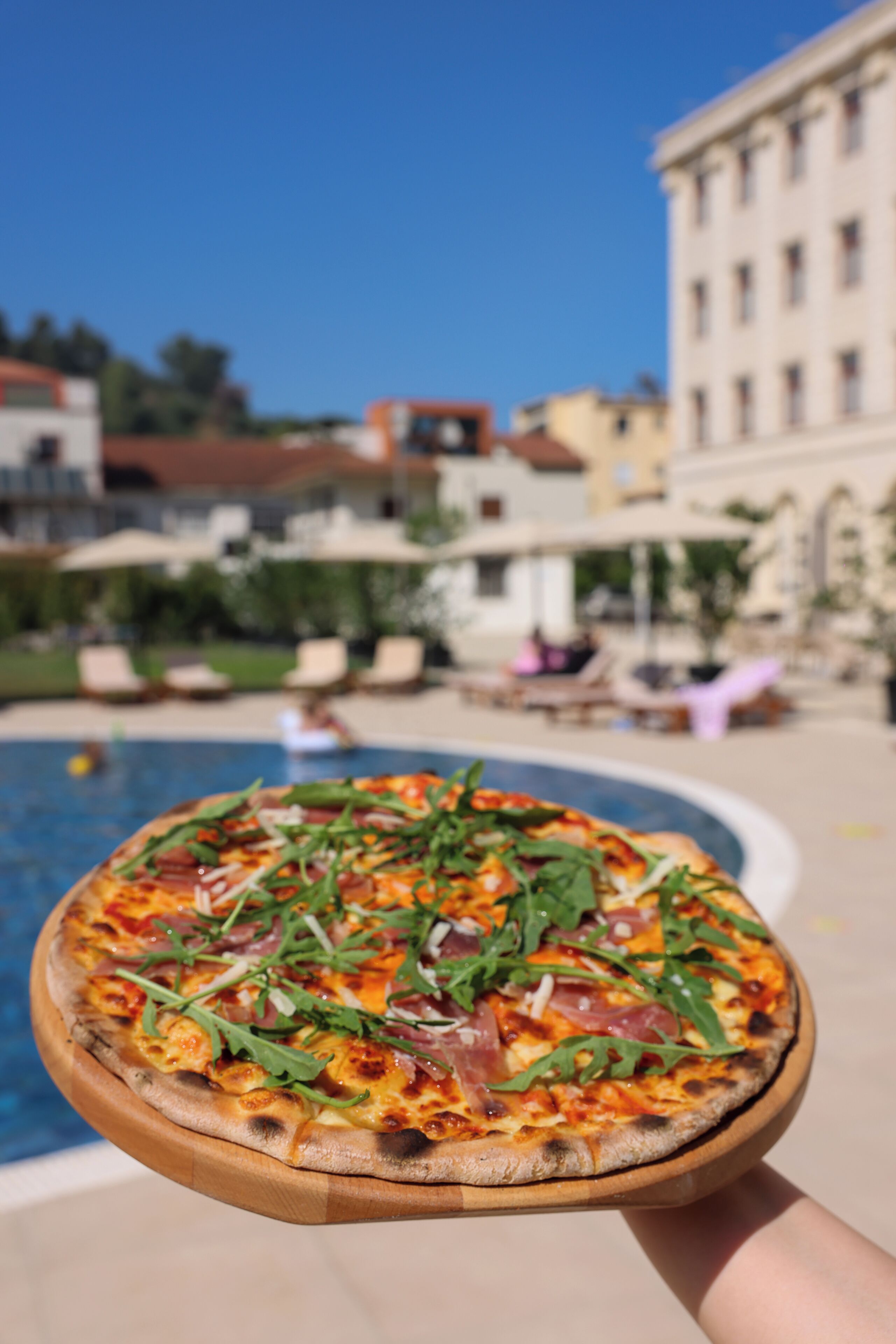Pizza com vegetais e queijo sobre uma tábua de madeira, exibida em frente a uma piscina com espreguiçadeiras e edifícios ao fundo.