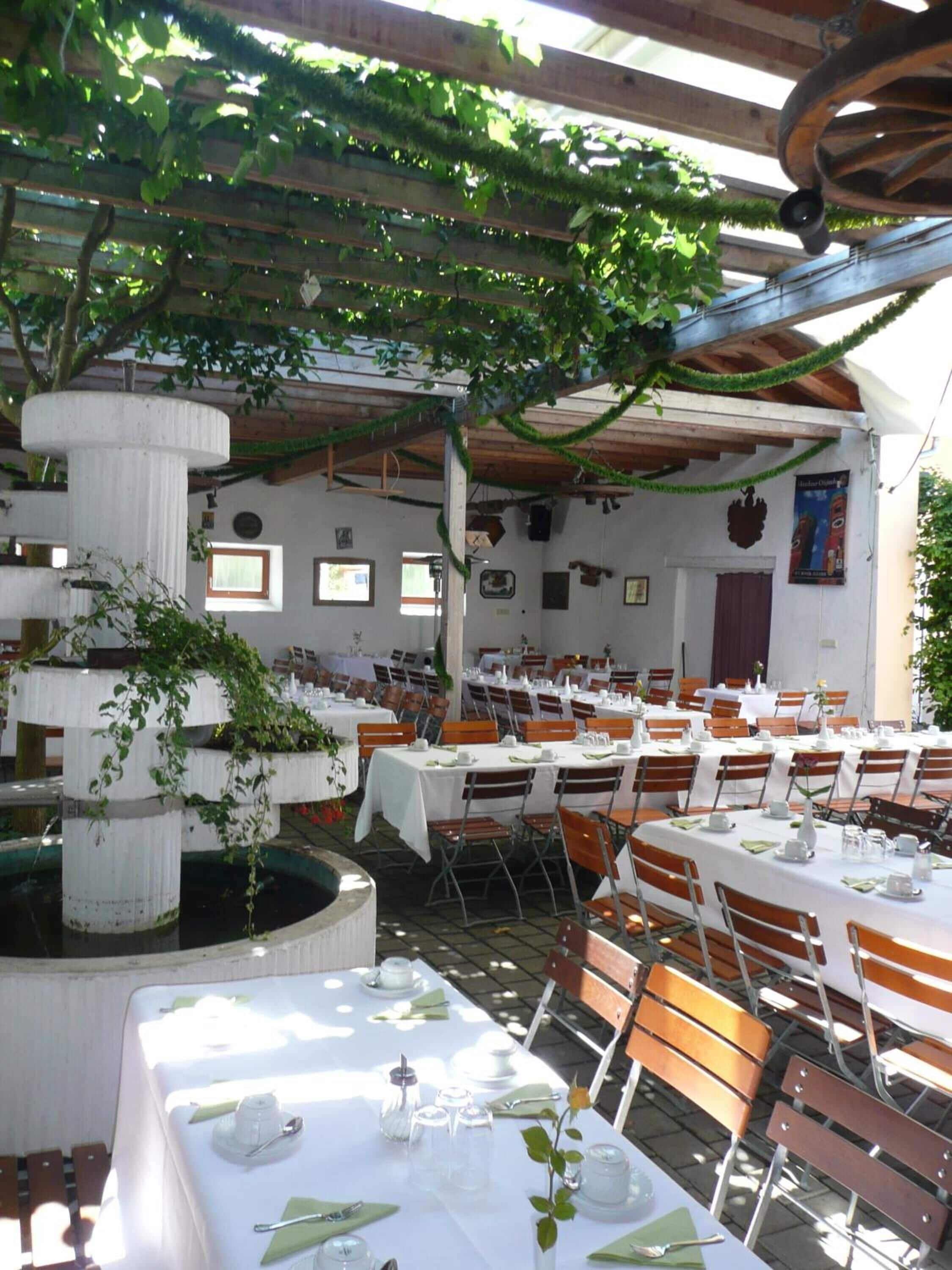 Uma área de refeições ao ar livre com mesas preparadas para o serviço, coberta por uma pérgola de madeira com trepadeiras, e uma fonte central.