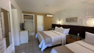 Quarto de hotel com duas camas, um guarda-roupa, uma televisão e uma parede de tijolos com um candeeiro.