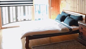 Quarto com teto de madeira, cama com almofadas azuis, mesa de cabeceira e vista para o exterior através de amplas janelas.