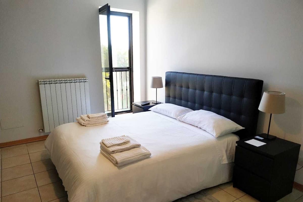 Uma cama bem arrumada com lençóis brancos e toalhas dobradas, uma cabeceira preta, uma mesa de cabeceira com abajur e um radiador na parede.