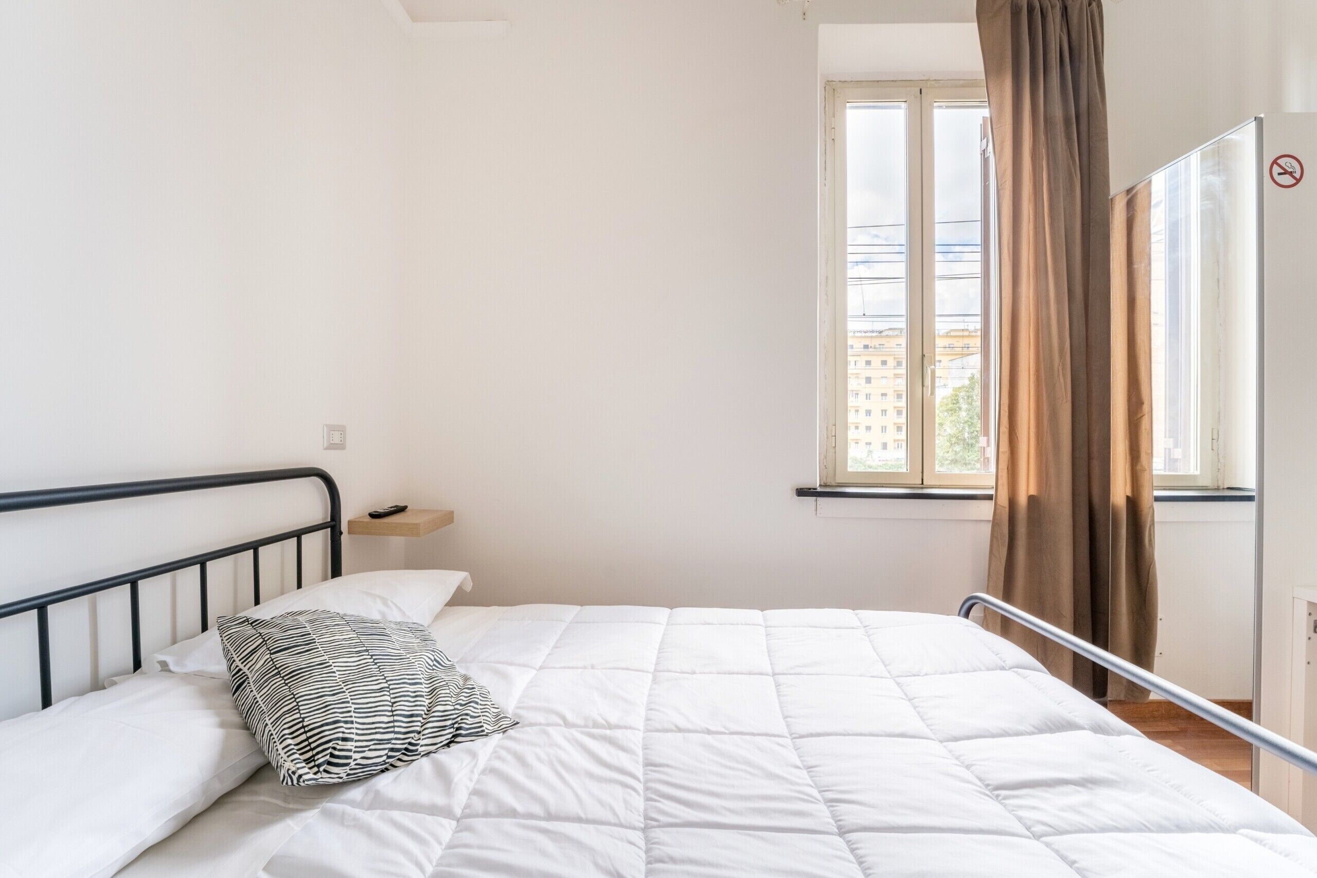 Uma cama de solteiro com colcha branca, travesseiro listrado e cabeceira de metal. Há uma janela com cortinas, uma mesa de cabeceira com abajur e uma porta com aviso de proibido fumar.