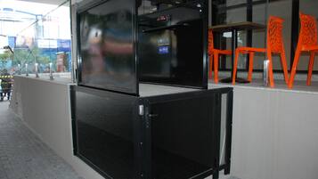 Estrutura de metal preto com painel de vidro, ao lado de um edifício com cadeiras laranja e uma mesa.