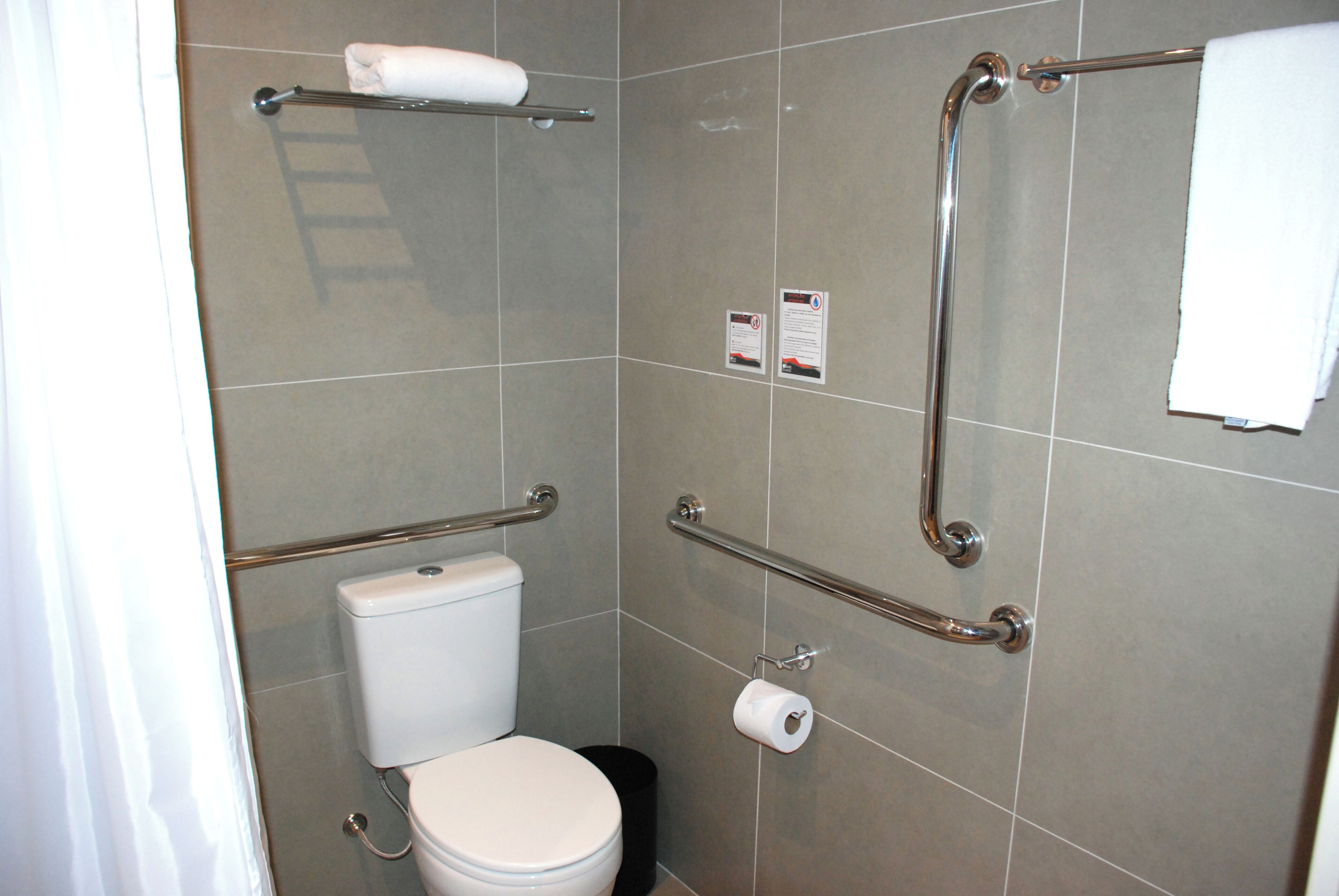 Uma casa de banho com sanita, barras de apoio, suporte para toalhas e rolo de papel higiénico.