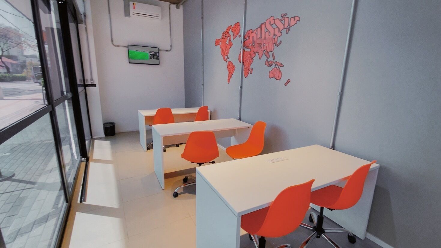 Um espaço de escritório moderno com mesas brancas e cadeiras laranja, um mapa-múndi na parede e uma janela com vista para o exterior.