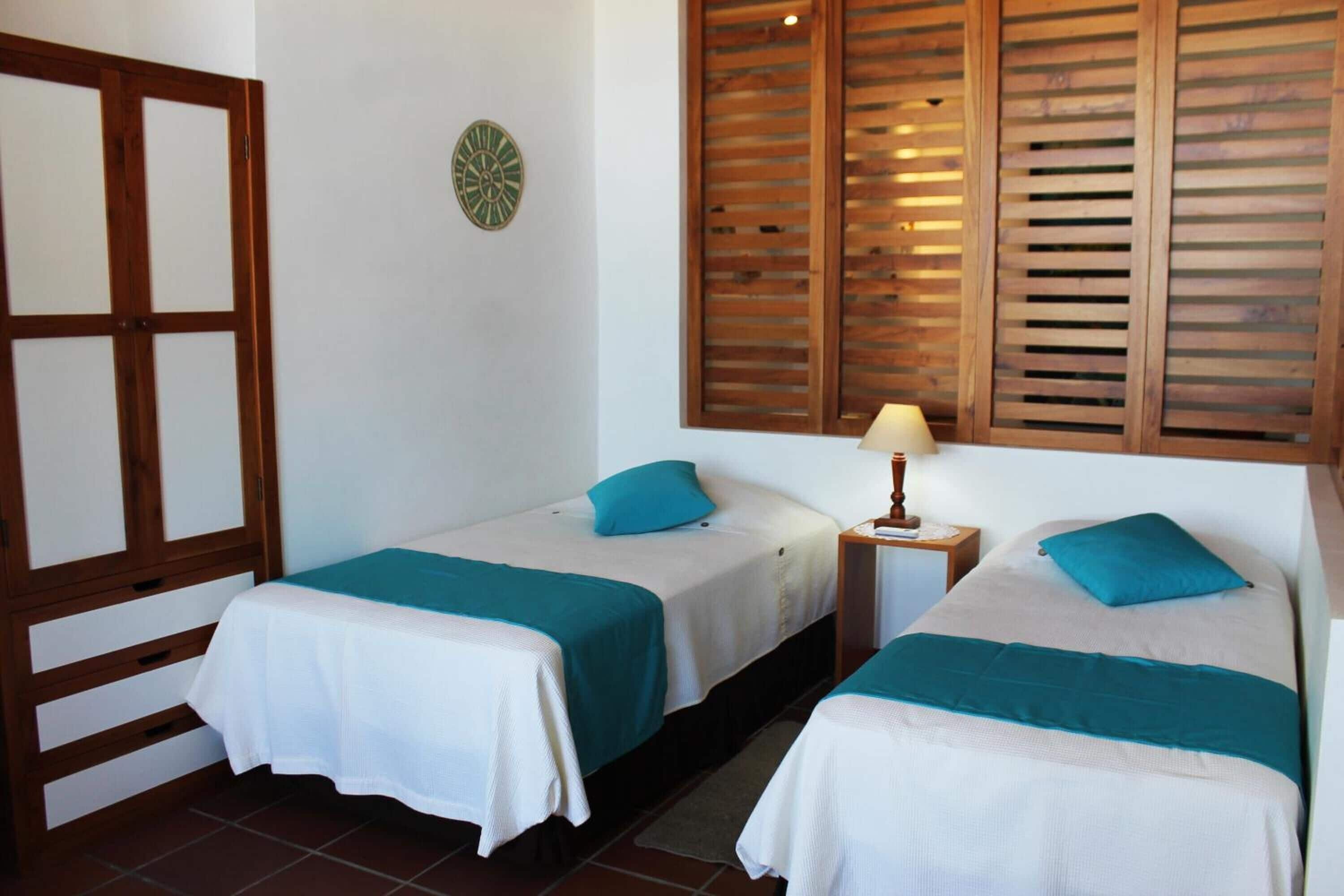Quarto com duas camas de solteiro, cada uma com almofada verde-água e roupa de cama branca. Possui um guarda-roupa de madeira com porta de vidro, uma mesa pequena com abajur e um relógio de parede.