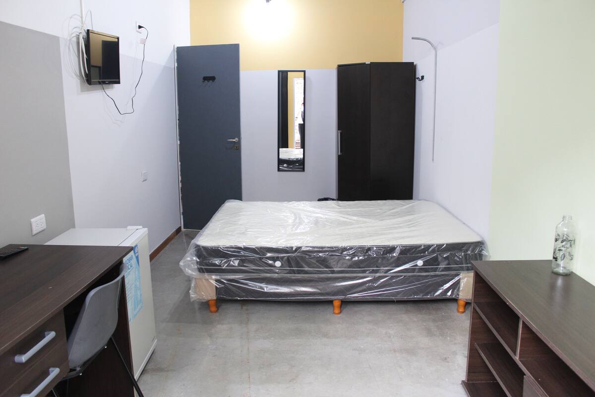 Quarto pequeno e limpo, com cama, escrivaninha e guarda-roupa.