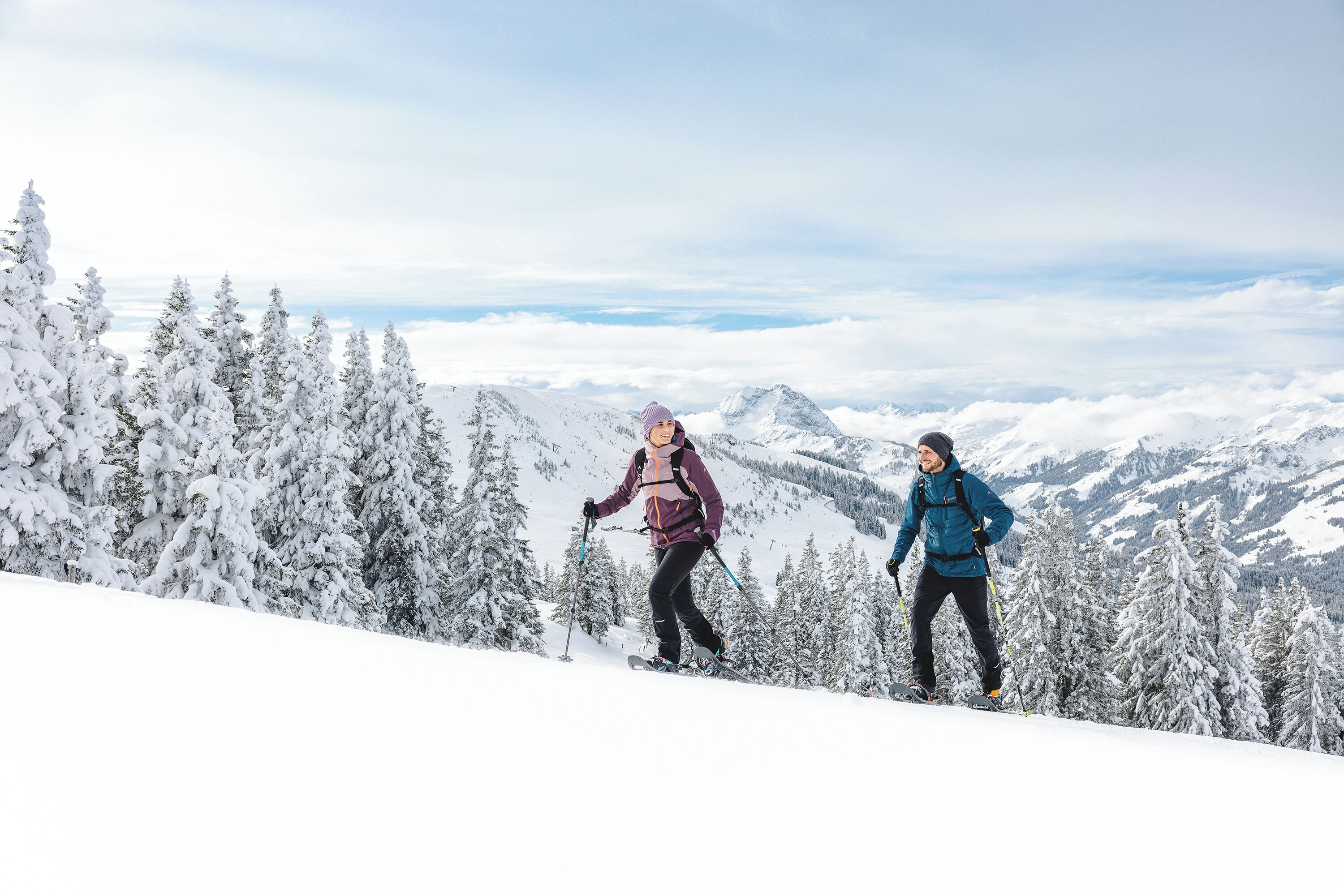 Duas pessoas esquiando numa montanha coberta de neve, com árvores e picos distantes ao fundo.