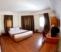 Quarto de hotel com uma cama grande, piso de madeira, uma escrivaninha com cadeira e cortinas nas janelas.