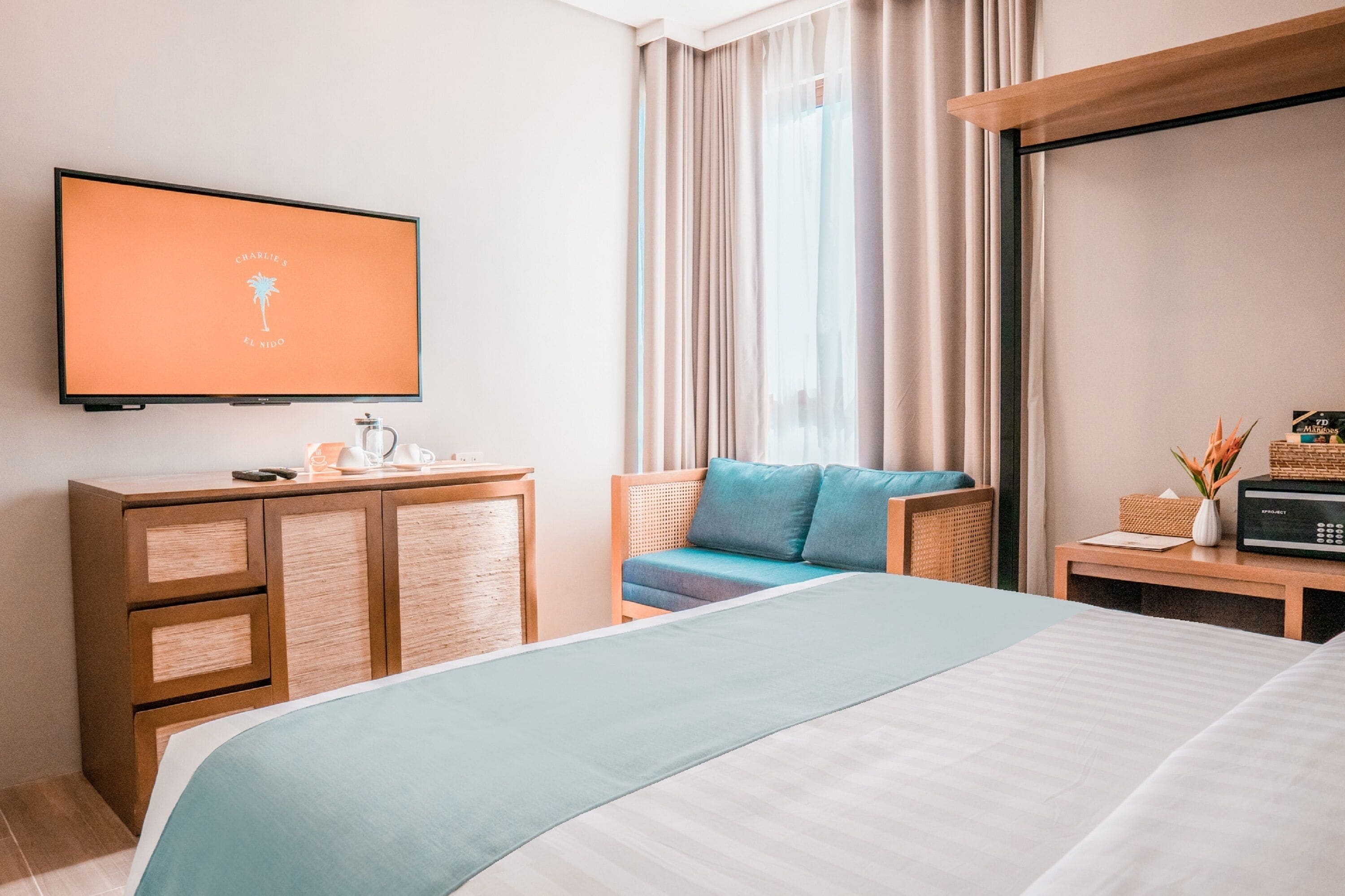 Quarto de hotel moderno com cama, TV fixada na parede, cómoda de madeira, cadeira de vime e mesa de cabeceira com rádio.