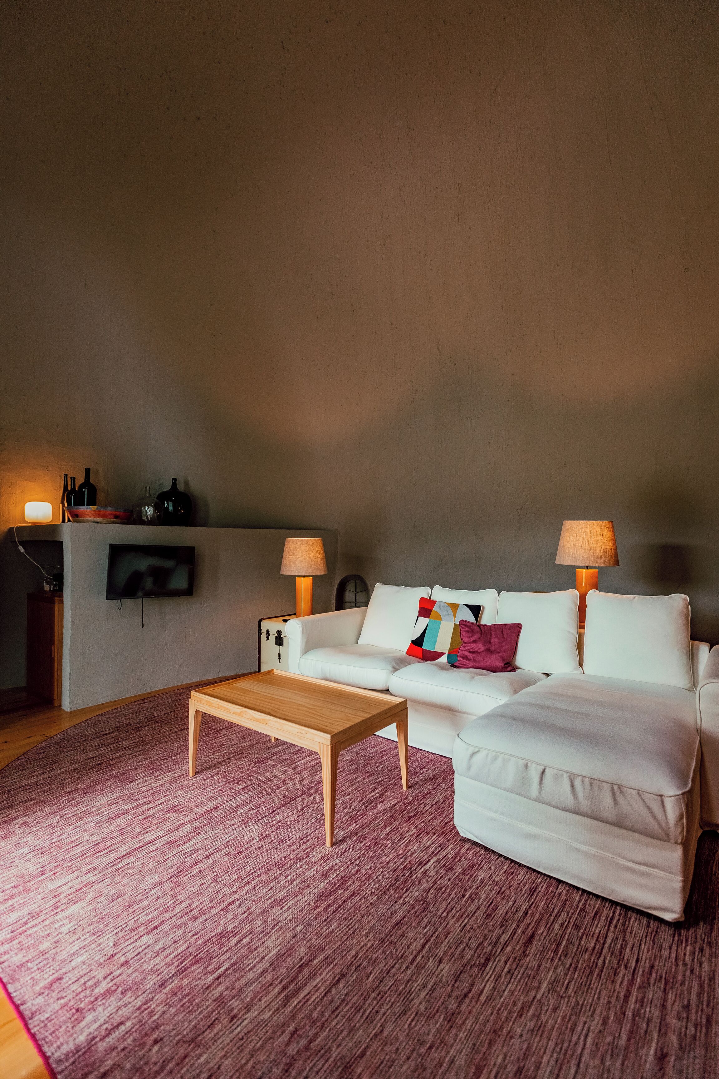 Uma sala de estar com um sofá branco, duas luminárias, uma mesa de centro de madeira e um tapete vermelho.