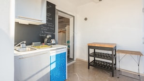 Uma cozinha compacta com uma lava-louças branca e azul, um quadro negro com anotações à mão e uma mesa de madeira com prateleiras.