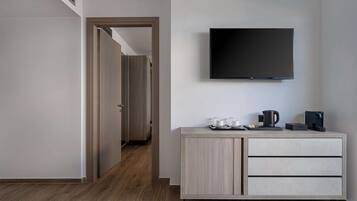 Quarto de hotel moderno com piso de madeira, televisão de tela plana na parede, frigobar com café e chá, e porta que dá acesso a outro quarto.