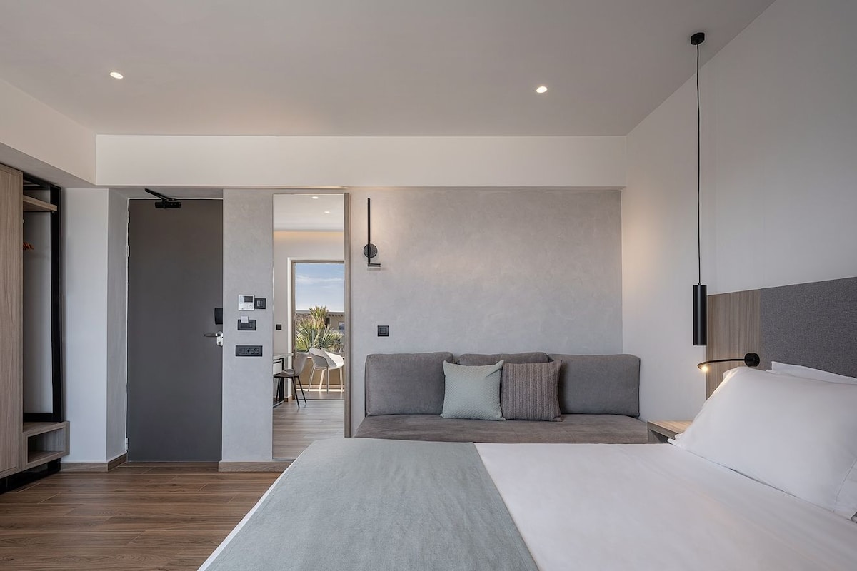 Quarto moderno com uma cama grande, um sofá e uma área de refeições visível através de uma porta aberta.