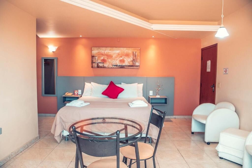Quarto de hotel com cama, mesa redonda com cadeiras, pequena escrivaninha e porta.