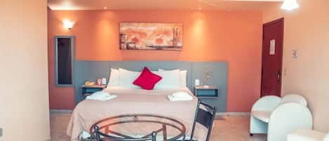 Quarto de hotel com cama, mesa redonda com cadeiras, pequena escrivaninha e porta.