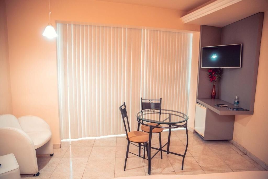 Quarto com mesa de vidro redonda, duas cadeiras, um sofá, televisão na parede e janela com persianas.