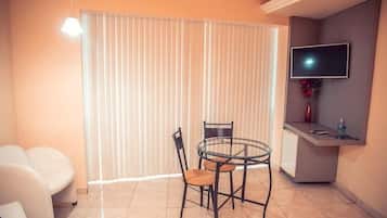 Quarto com mesa de vidro redonda, duas cadeiras, um sofá, televisão na parede e janela com persianas.