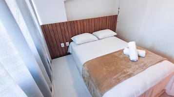 Quarto de hotel com cama, cabeceira de madeira, travesseiro branco, manta marrom e toalha enrolada na cama.
