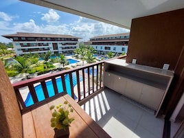 Uma varanda com vista para uma piscina, área de refeições com mesa e cadeiras, e um edifício com vários andares e varandas.