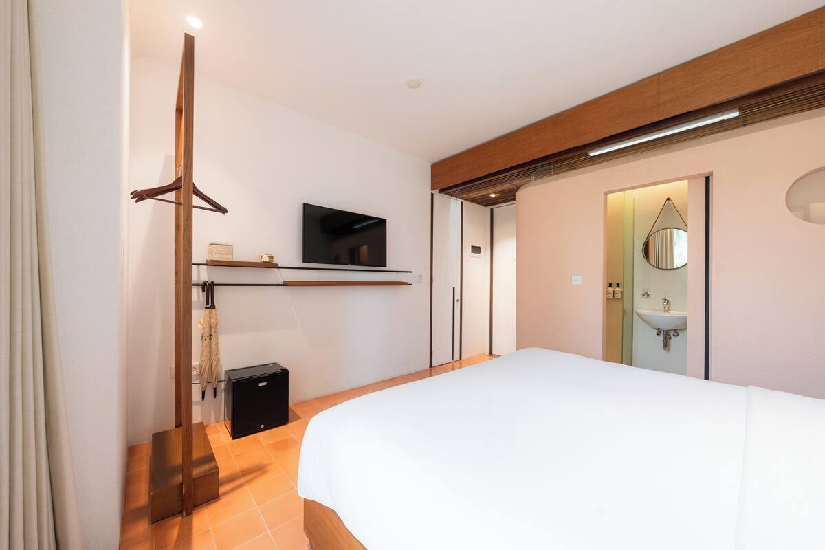 Quarto de hotel moderno com uma cama grande, uma televisão de tela plana na parede, um cabide de madeira, um pequeno armário e um banheiro visível através de uma porta aberta.