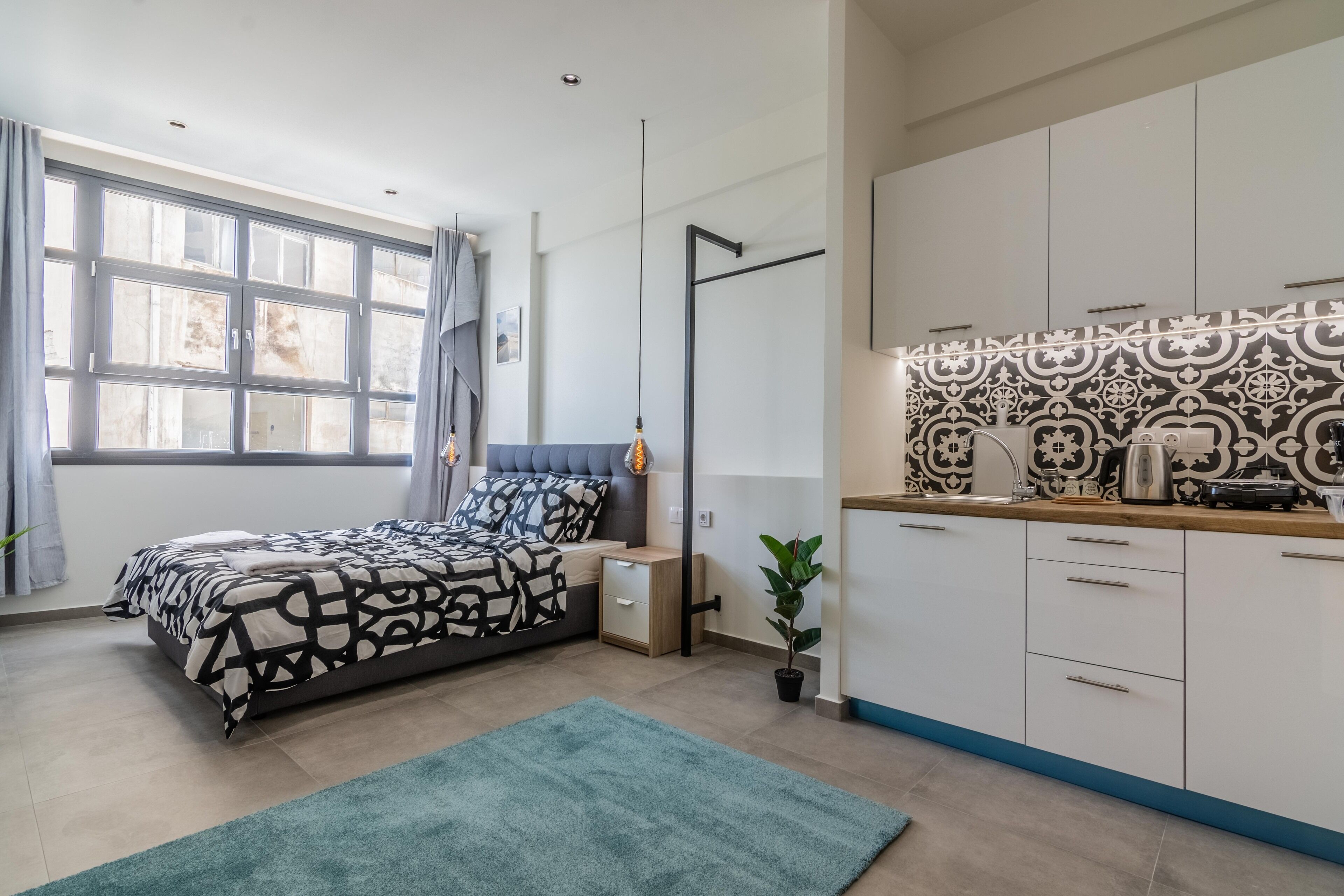 Um quarto moderno com uma cama grande, mesas de cabeceira, um criado-mudo, uma planta em vaso e vista para o exterior através de uma janela com cortinas.