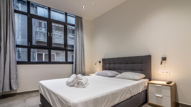 Cama bem arrumada com colcha branca e duas almofadas, mesa de cabeceira com abajur e janela com cortinas.