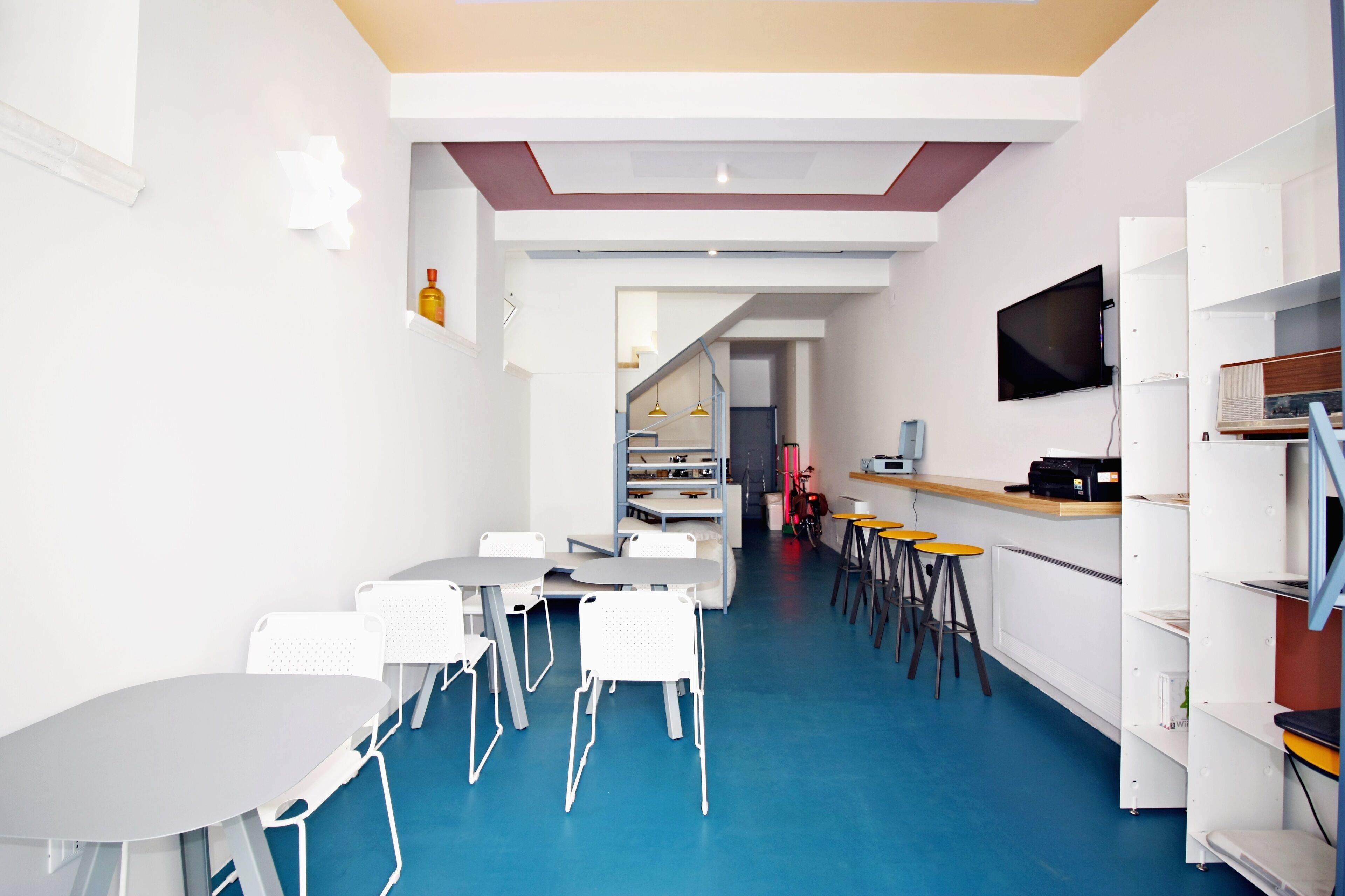 Quarto moderno e bem iluminado, com paredes brancas, chão azul e área de cozinha com televisão e bancada.
