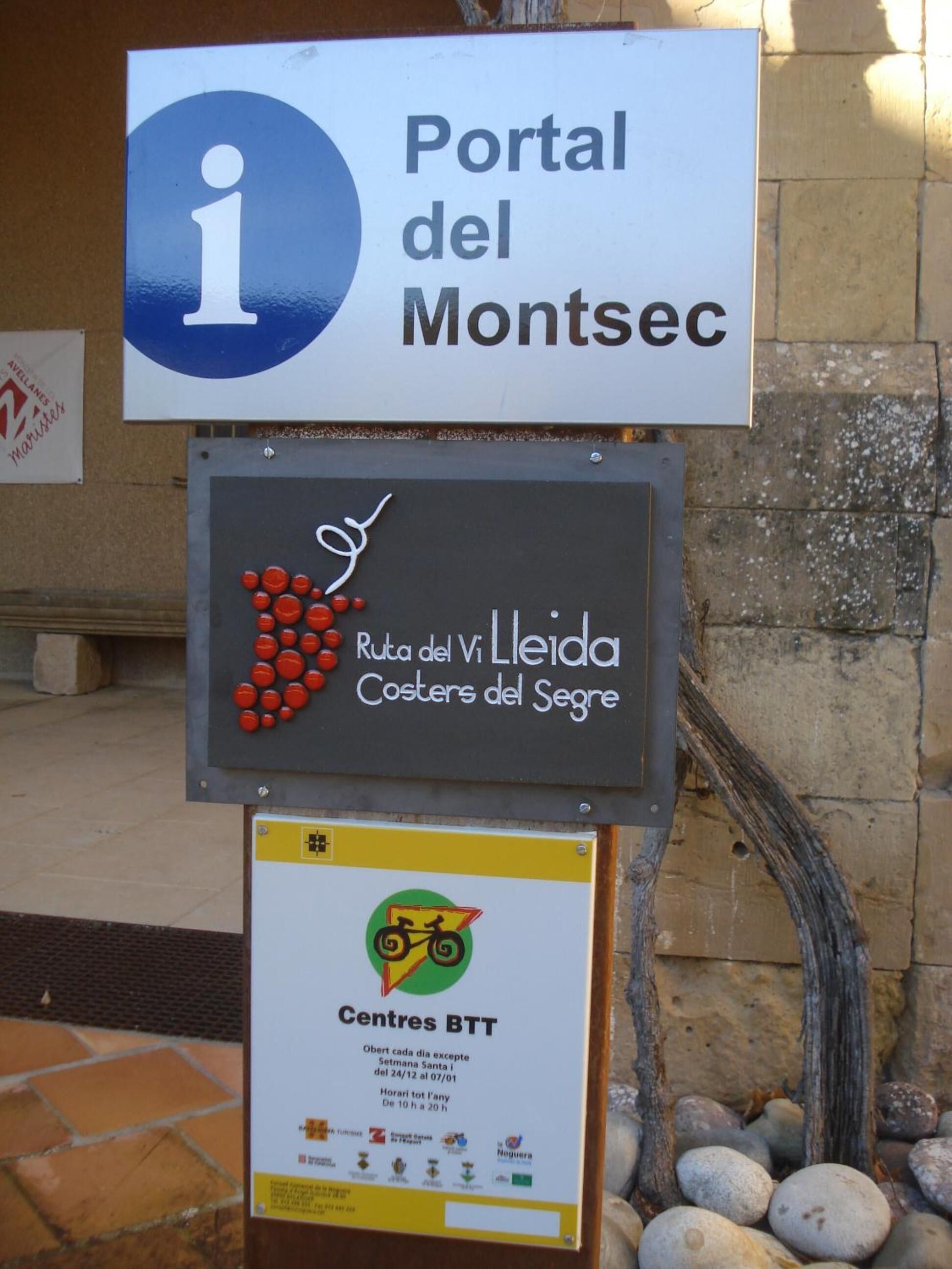 Um sinal para 'Portal del Montsec' com o símbolo de uma montanha e um círculo vermelho. Abaixo, um sinal para 'Ruta del Vi Lleida Costers del Segre' com um círculo vermelho e uma linha branca. Na parte inferior, um sinal para 'Centres BTT' com o símbolo de uma bicicleta e informações sobre horários de funcionamento.