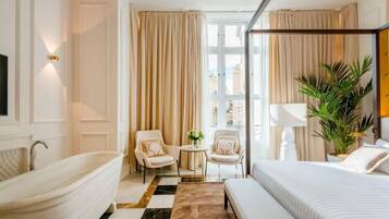 Quarto de hotel com uma cama grande, área de estar com duas cadeiras, uma mesinha com um vaso, banheira e uma janela ampla com cortinas.