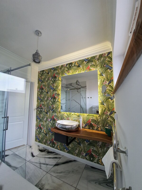 Casa de banho com chão em azulejos, lavatório com espelho e parede com papel de parede de pássaros tropicais.