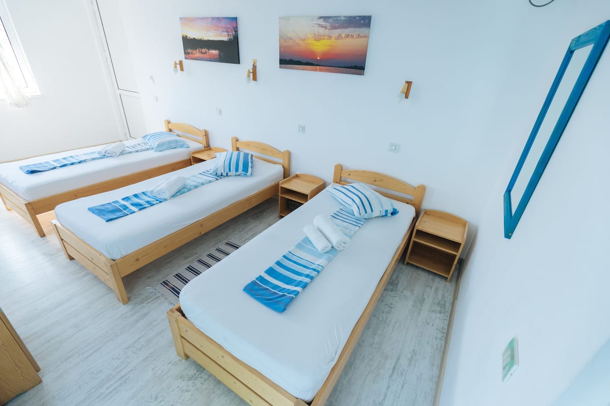 Quarto com três camas de madeira, cada uma com colchão branco e roupa de cama listrada em azul. Há duas pinturas nas paredes retratando paisagens.