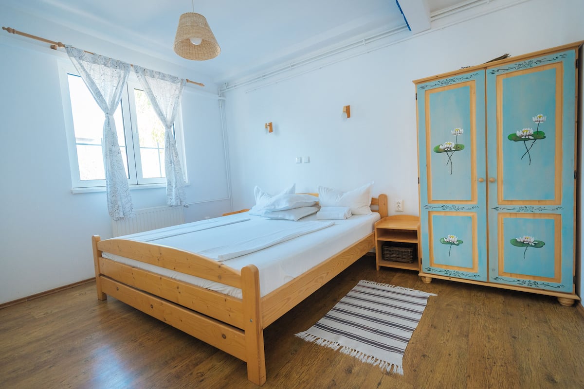Quarto com cama de madeira, guarda-roupa com estampas florais, janela com cortinas leves e chão de madeira.