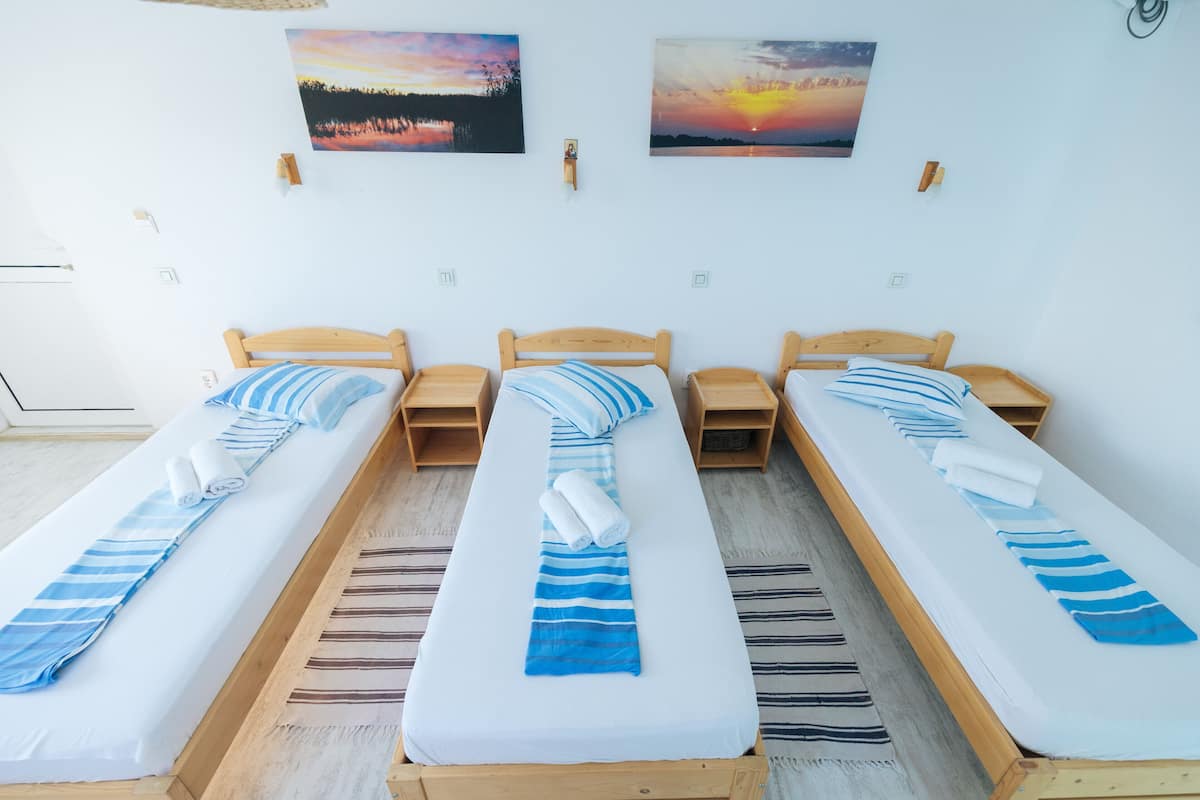 Quarto de hotel com três camas simples, cada uma com cabeceira de madeira, roupa de cama branca e toalhas com listras azuis.