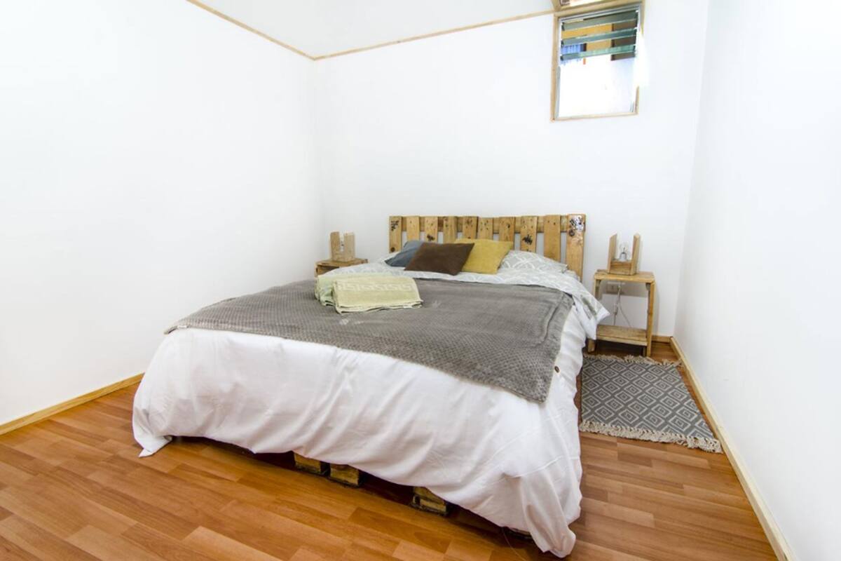 Quarto com cama de madeira, roupa de cama branca, uma pequena mesa de cabeceira com abajur e um tapete estampado.