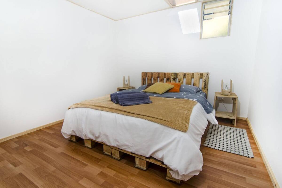 Quarto com estrutura de cama em madeira, roupa de cama branca e uma pequena mesa de cabeceira de madeira com abajur.