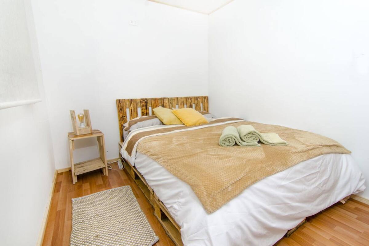 Quarto com cabeceira de madeira, cama com roupa de cama branca e bege, duas almofadas e uma pequena mesa de cabeceira de madeira com abajur.