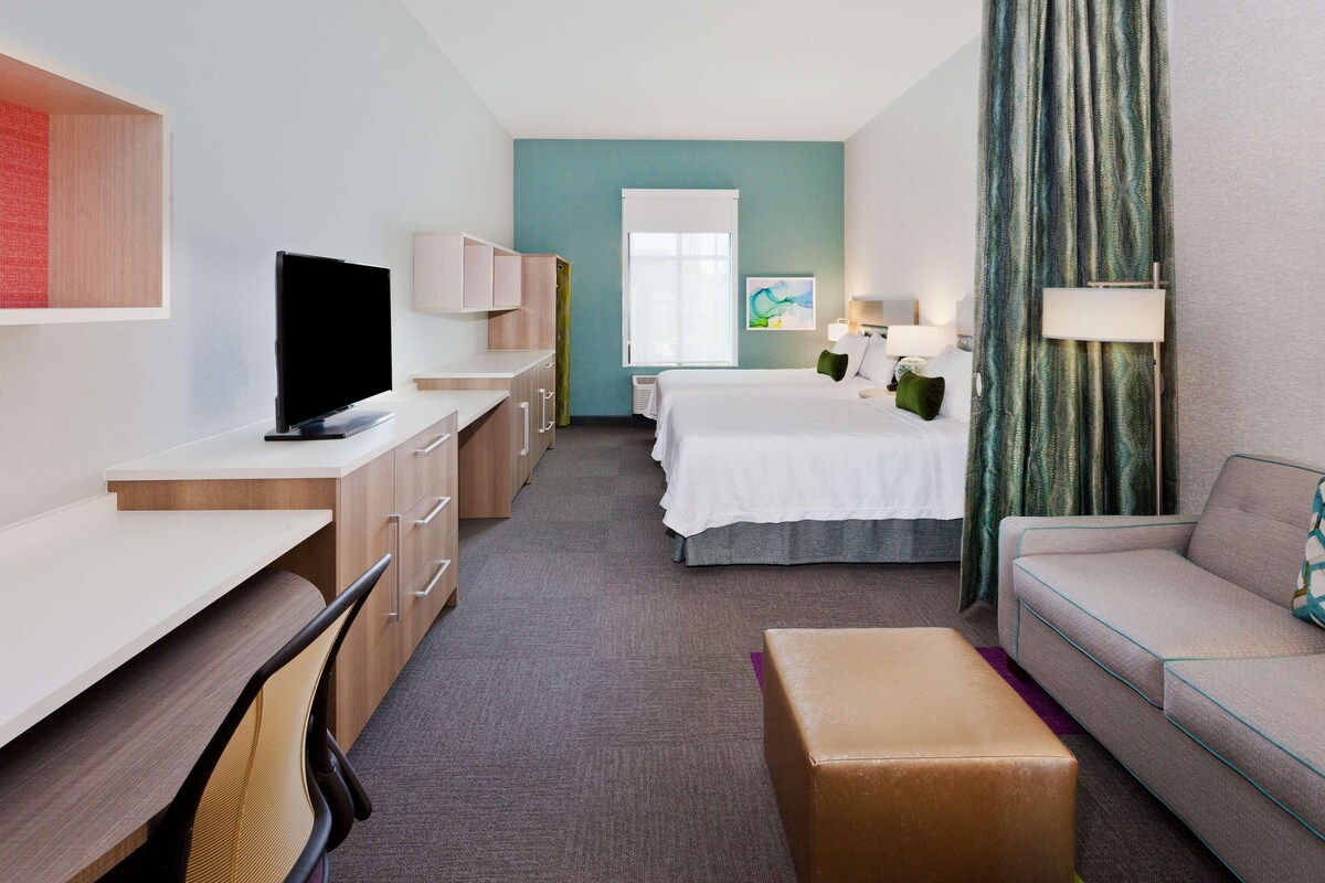 Quarto de hotel com cama, sofá, escrivaninha com televisão e janela com cortinas.
