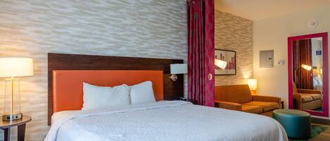 Quarto de hotel com uma cama grande, cabeceira de madeira, mesa de cabeceira com abajur, área de estar com poltrona e banqueta, e espelho com moldura vermelha.