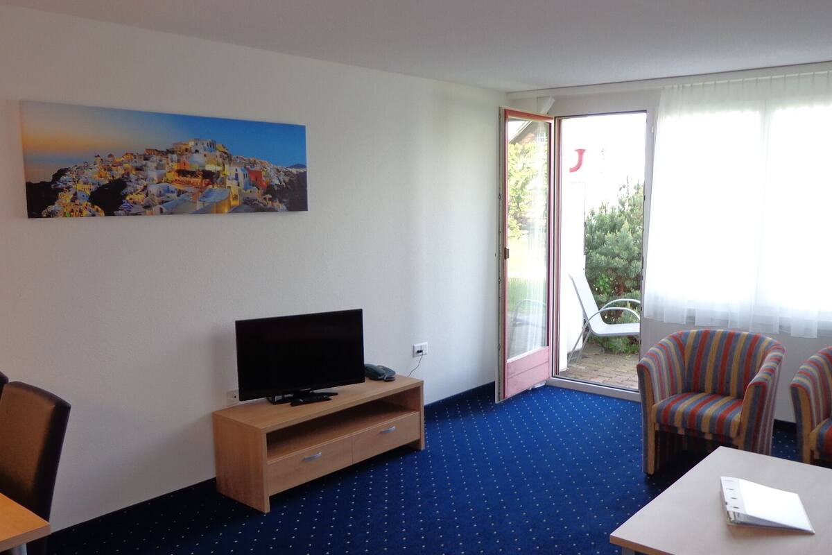 Quarto de hotel com televisão sobre suporte de madeira, poltronas listradas, uma mesinha e um quadro de uma paisagem costeira.