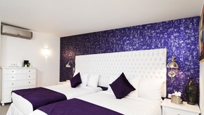 Quarto de hotel com duas camas, uma cômoda branca, uma mesinha de cabeceira e uma parede roxa com textura.
